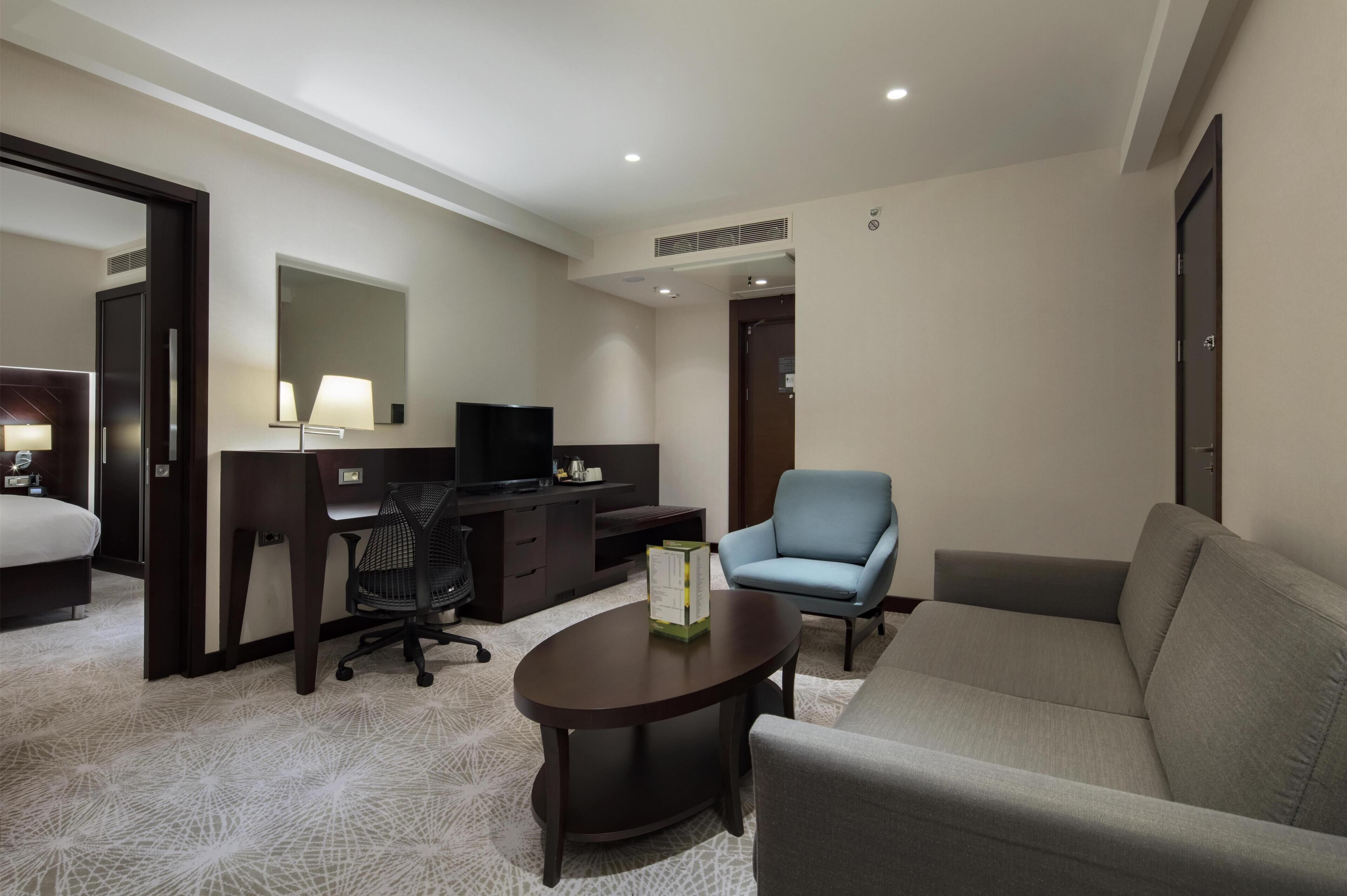 Quarto de hotel com cama, escrivaninha com cadeira, sofá, mesa de centro e televisão.