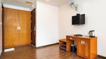 Quarto com guarda-roupa de madeira, pequena escrivaninha com chaleira, lixeira e televisão presa à parede.
