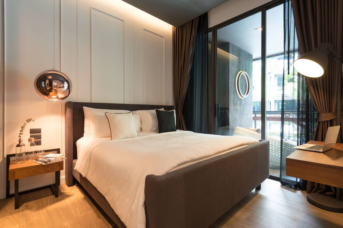 Quarto de hotel moderno com uma cama grande, uma escrivaninha com um laptop, uma mesinha de cabeceira com uma luminária e vista para o exterior através de uma janela ampla.