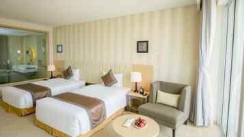 Quarto de hotel com duas camas, um sofá, uma mesa redonda com frutas, um espelho e uma janela com cortinas.