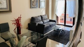 Uma sala de estar moderna com um sofá de couro preto, uma mesa de centro de vidro e um espaço de jantar com mesa e cadeiras de vidro. Há duas obras de arte emolduradas na parede e uma janela com cortinas leves.