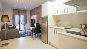 Uma cozinha moderna com armários brancos, uma geladeira de aço inoxidável e uma pia. Há uma área de refeições com uma mesa e cadeiras, e um sofá ao fundo.