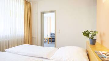 Quarto de hotel com duas camas, uma mesa de cabeceira de madeira com uma revista, uma planta em vaso e vista para uma sala de estar através de uma porta.