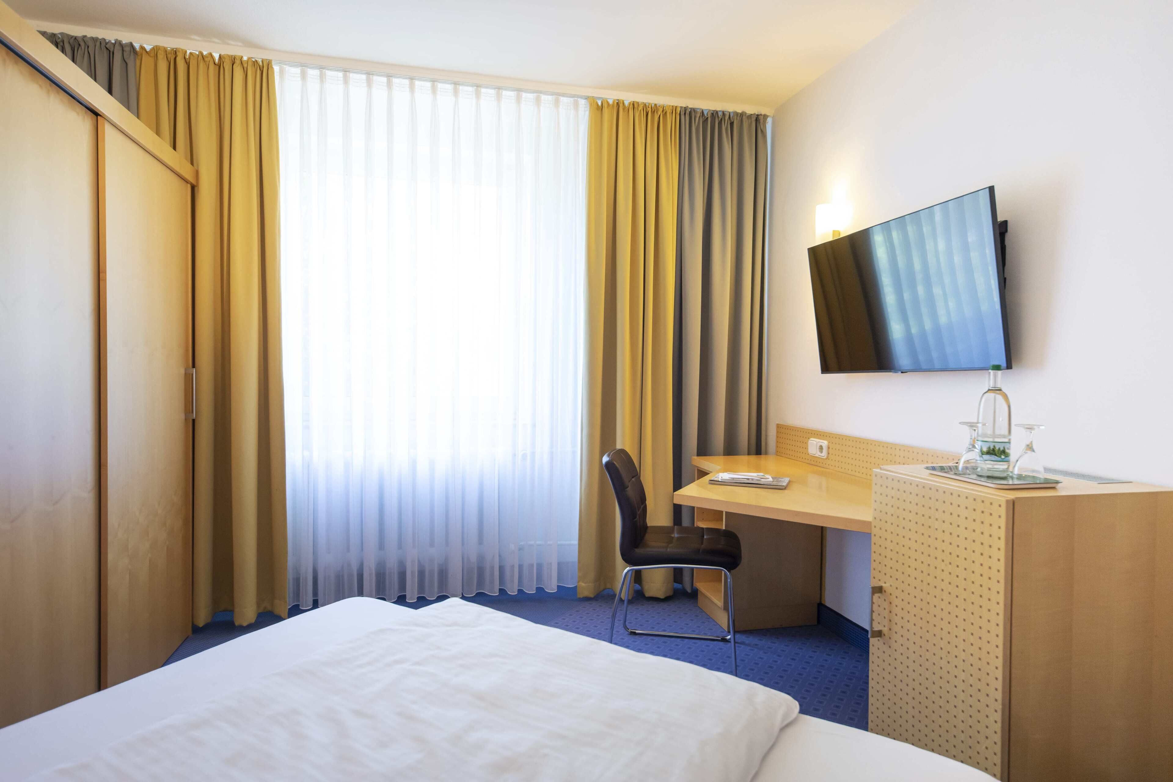 Quarto de hotel com cama, escrivaninha, cadeira, televisão e janela com cortinas.