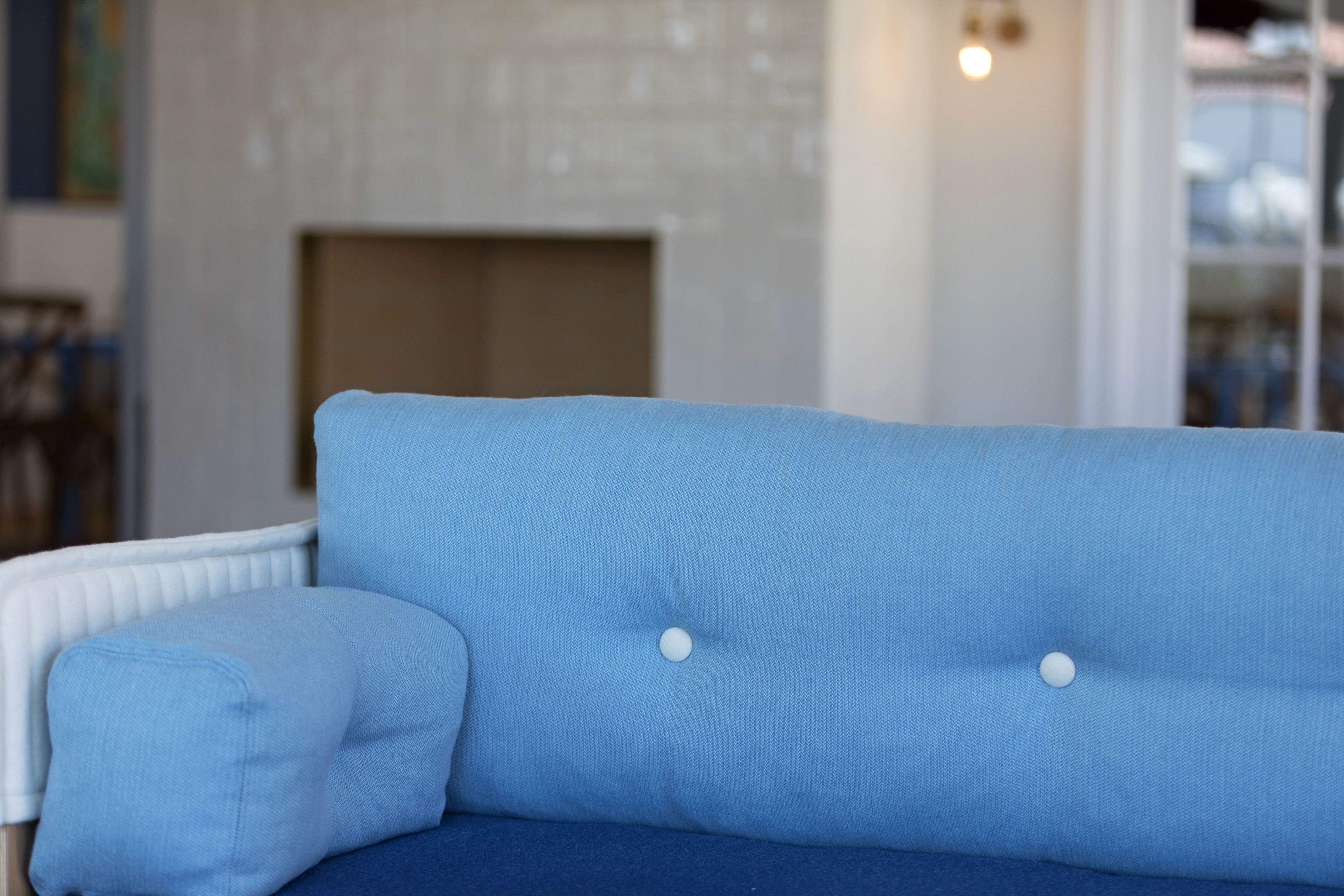 Um sofá azul com botões brancos, um travesseiro branco e uma cadeira branca num quarto com lareira e estante de livros.