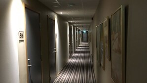 Corredor de hotel com carpete listrado, portas em ambos os lados e quadros nas paredes.