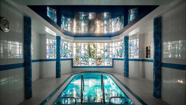 Piscina com água cristalina, rodeada por paredes azulejadas em azul e uma janela de vitrais.