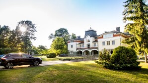 Uma casa grande, com vários andares e uma cúpula imponente no telhado, rodeada por jardins bem cuidados e árvores.