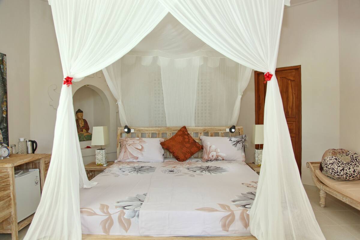 Quarto com cama de dossel, cabeceira de madeira, uma estátua e uma pequena mesa de cabeceira com abajur.