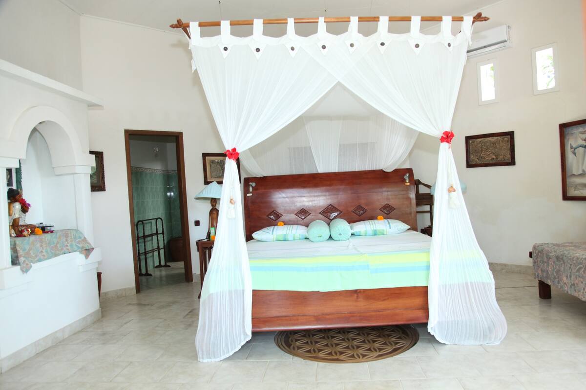 Quarto com cama de madeira, dossel branco e uma pequena mesa redonda com abajur.