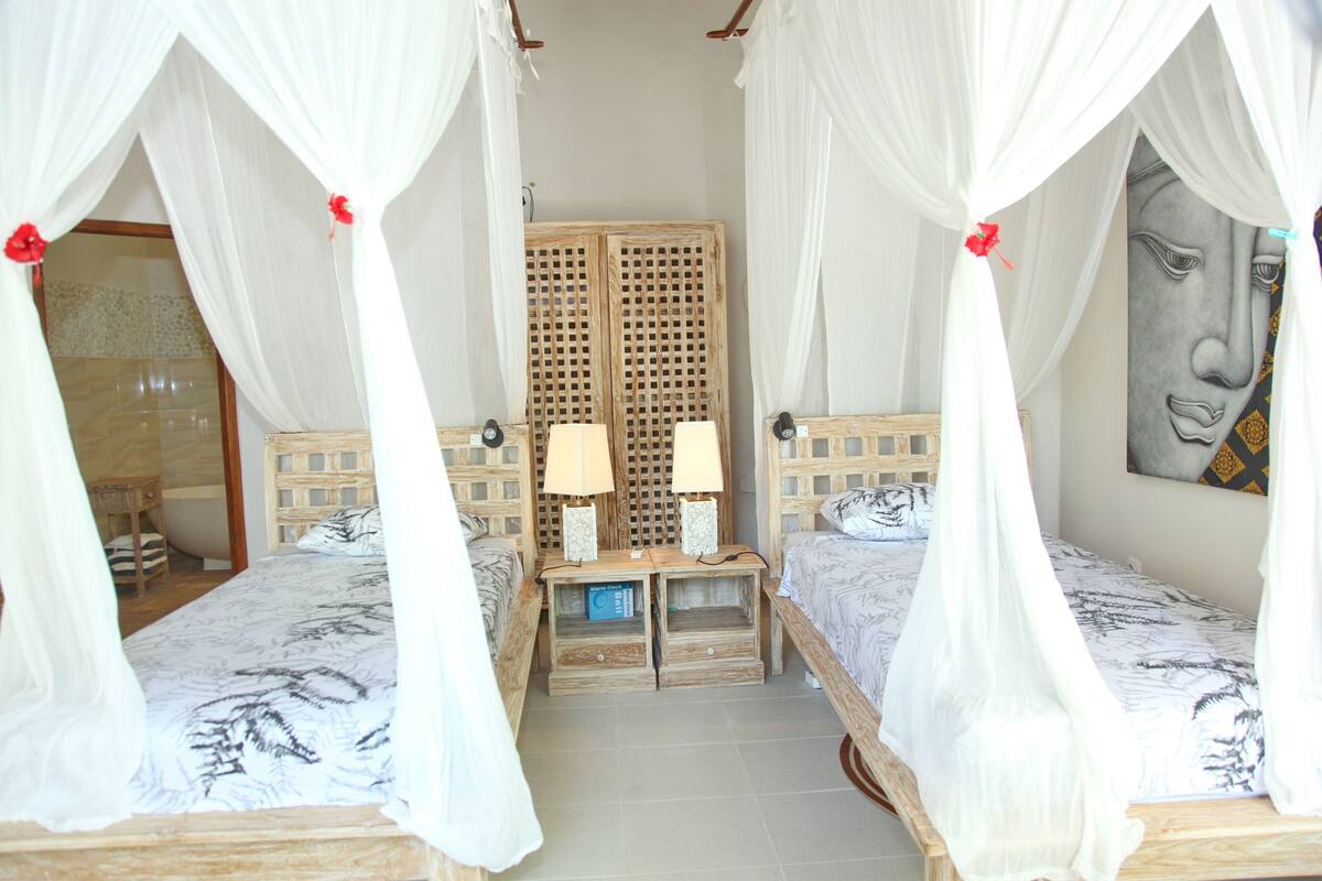 Quarto com duas camas, cada uma com dossel e cabeceira de madeira. Há mesas de cabeceira com abajures e uma pequena estante entre as camas.