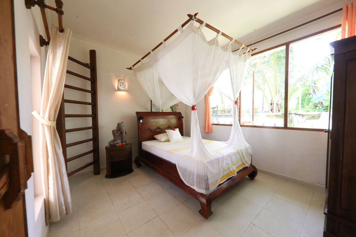 Quarto com cama de madeira, dossel branco, mesa de cabeceira, escada de madeira e vista para o verde lá fora.