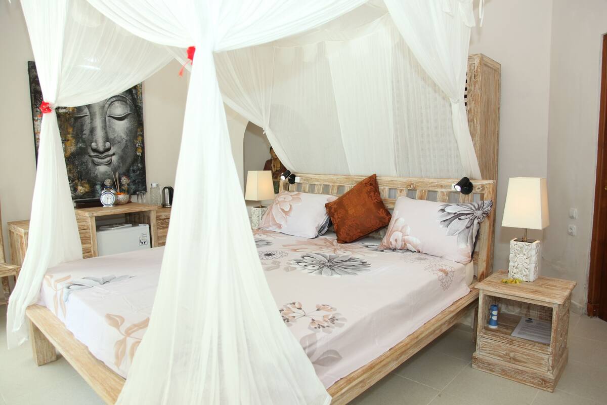 Quarto com cama de dossel, cabeceira de madeira, mesas de cabeceira com luminárias e uma obra de arte na parede.
