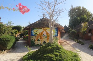 Uma construção com telhado de palha e um mural de girafa, cercada por vegetação e um caminho de cascalho.
