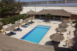 Uma área de piscina com espreguiçadeiras e guarda-sóis de palha, cercada por um edifício com grandes janelas.