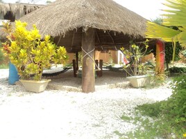 Uma cabana com telhado de palha e chão de cascalho, cercada por vasos de plantas e uma rede.