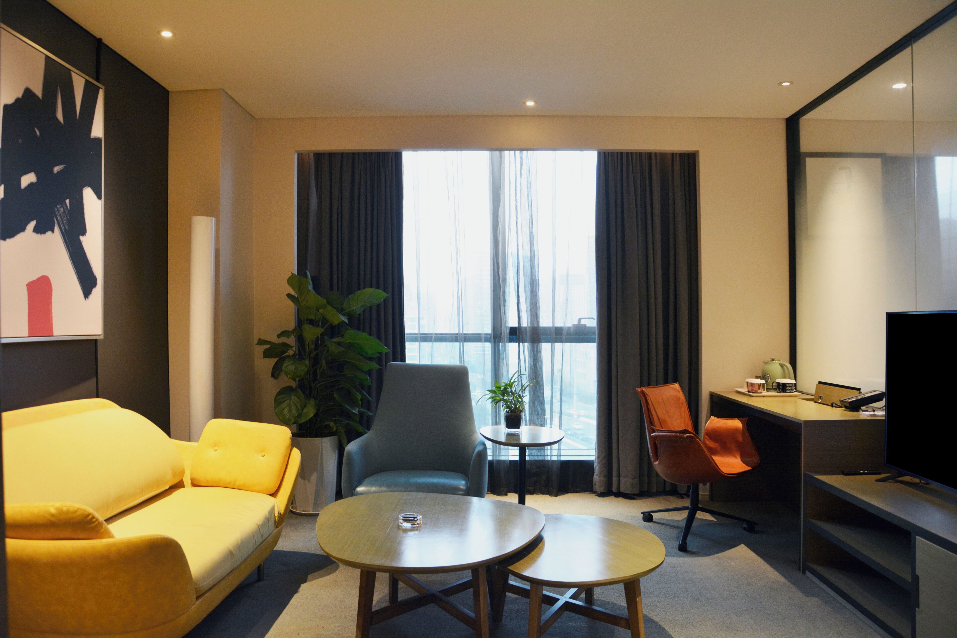 Quarto de hotel moderno com um sofá amarelo, duas mesas redondas, uma grande planta em vaso, uma escrivaninha com televisão e uma janela grande com cortinas.