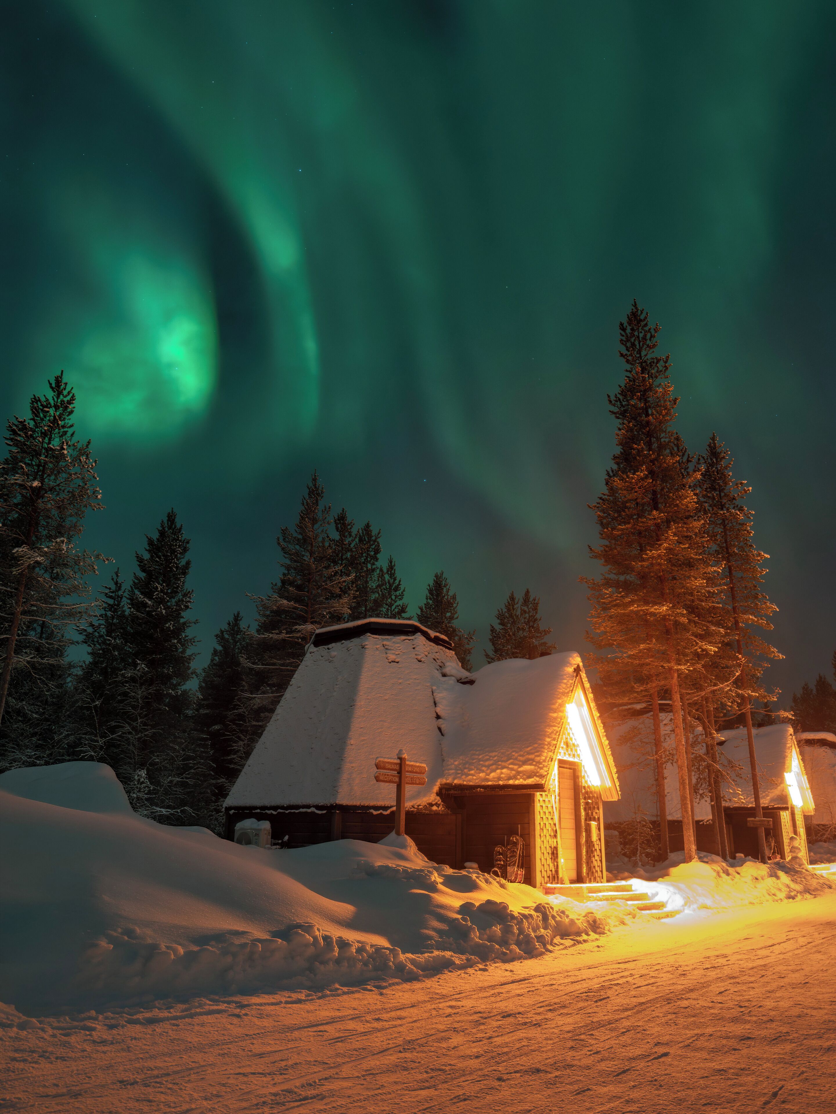 Uma cabana coberta de neve com telhado de madeira, rodeada por árvores, sob um céu noturno com a aurora boreal.