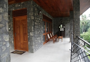 Um edifício de pedra com porta de madeira, um alpendre coberto com cadeiras e vista para o verde.
