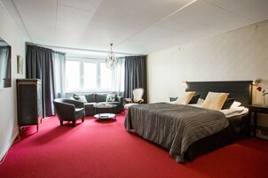 Quarto de hotel com uma cama grande, um sofá, uma poltrona, uma mesinha e uma escrivaninha com uma luminária.