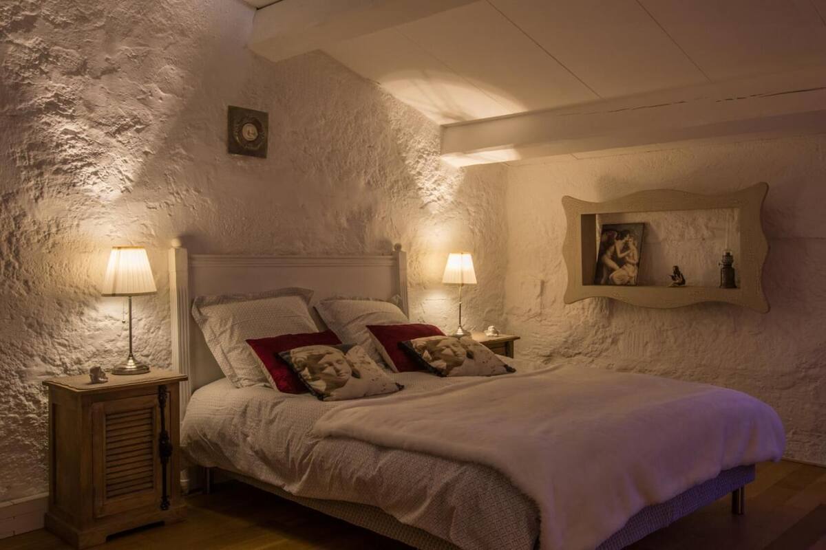 Um quarto com parede de pedra, estrutura de cama de madeira, duas mesas de cabeceira com abajures e uma prateleira fixada na parede com objetos decorativos.