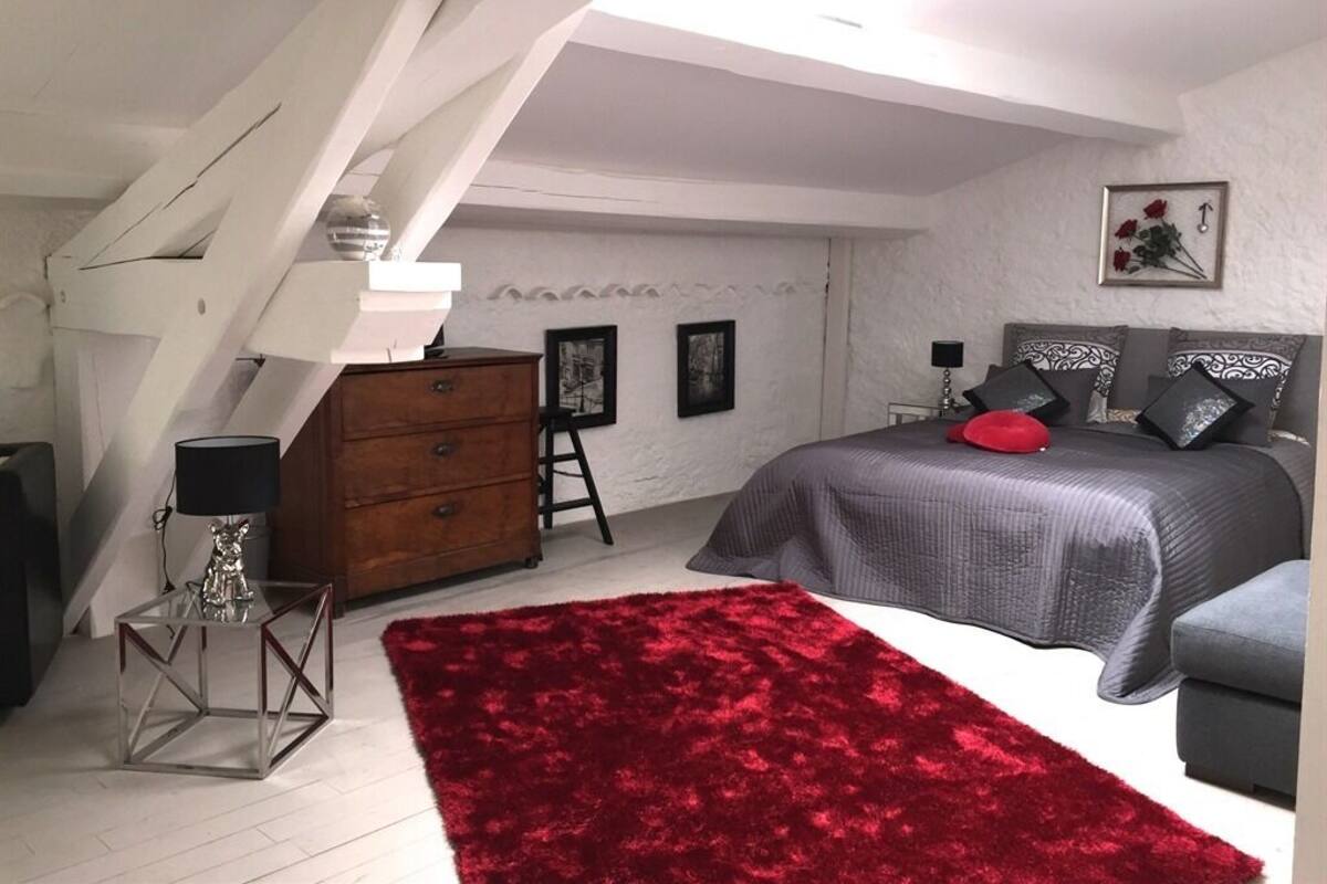 Quarto com teto inclinado, cama com roupa de cama cinza, tapete vermelho, cómoda de madeira e quadro emoldurado na parede.