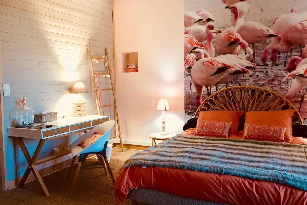 Quarto com escrivaninha de madeira, uma cadeira, cama com roupa de cama laranja e cinza, e um mural de flamingos na parede.