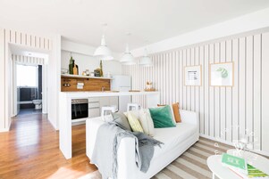Uma sala de estar moderna com sofá branco, tapete listrado e área de cozinha com armários de madeira e iluminação pendente.