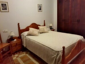 Uma cama bem arrumada com cabeceira de madeira, mesa de cabeceira com abajur e um tapete xadrez no chão.