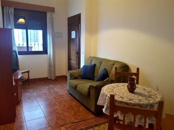Uma sala de estar com um sofá, uma mesa pequena com toalha de renda, uma televisão e uma janela com cortinas.