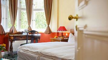 Quarto de hotel com cama, escrivaninha, cadeira, abajur e janela com cortinas.