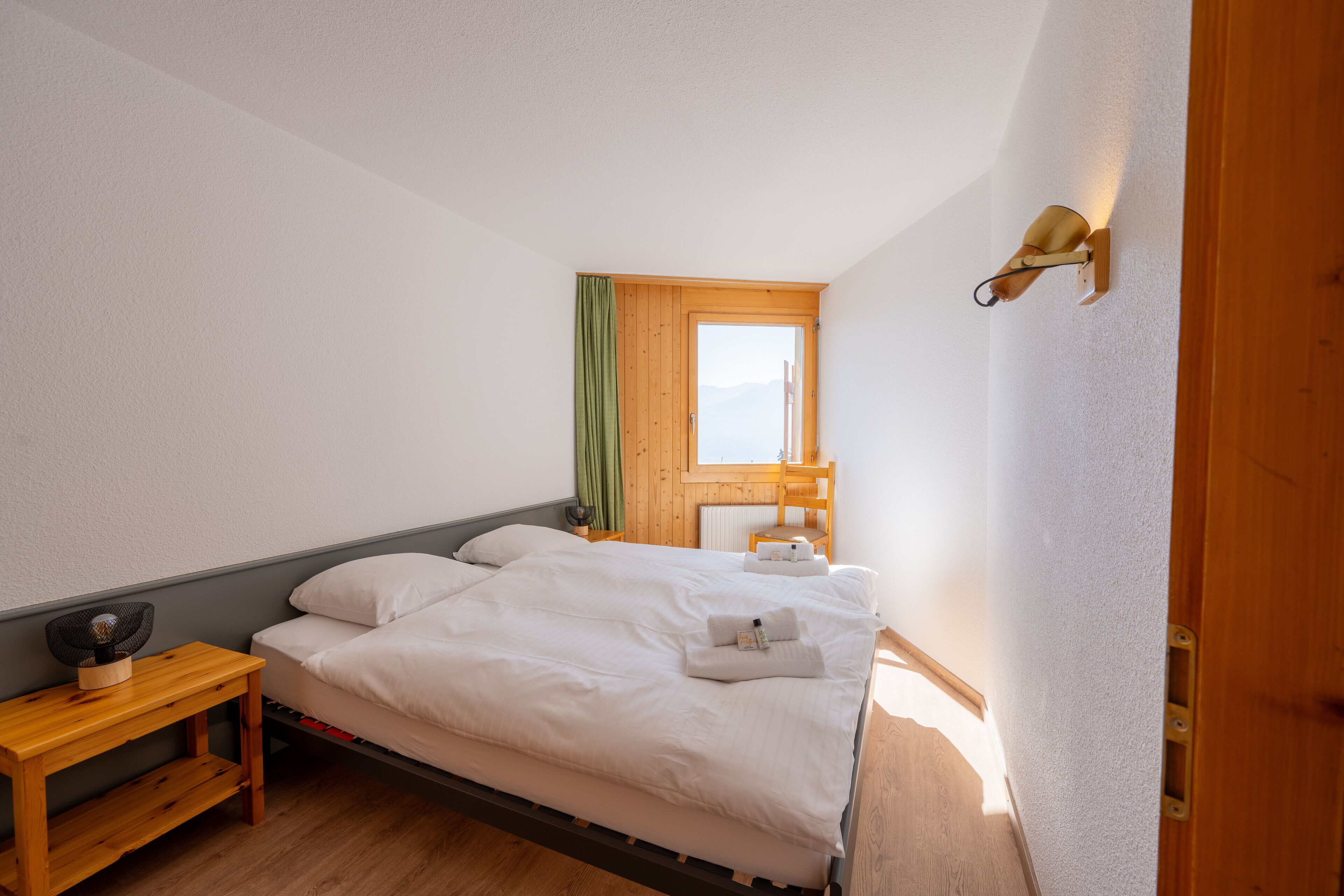 Quarto com cama, mesa de cabeceira, janela com cortinas e luminária na parede.