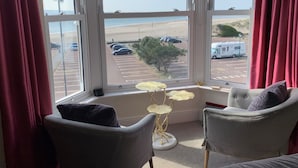 Quarto com uma grande janela com vista para uma praia e estacionamento, com duas cadeiras e uma pequena mesa.