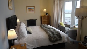 Quarto de hotel com cama, luminárias de cabeceira, uma cadeira, escrivaninha, janela com cortinas e vista para o mar.