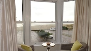 Quarto com janela em sacada com vista para a praia, duas poltronas, uma mesa pequena e um veículo estacionado na praia.