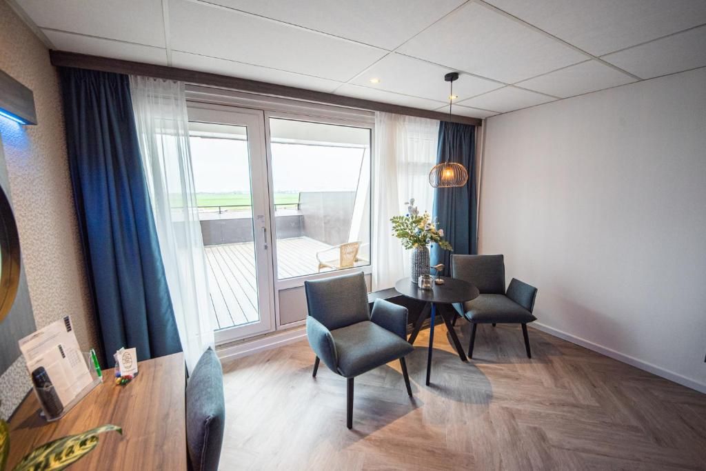 Quarto de hotel moderno com piso de madeira, mesa de jantar com cadeiras, uma grande janela com cortinas e luminária pendente.