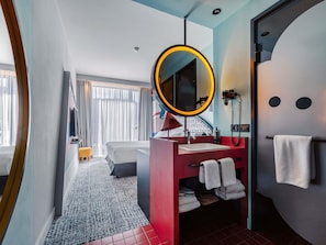 Quarto de hotel com um espelho grande, uma toucador vermelha, uma pia branca, um porta-toalhas, uma cama e uma janela com cortinas.
