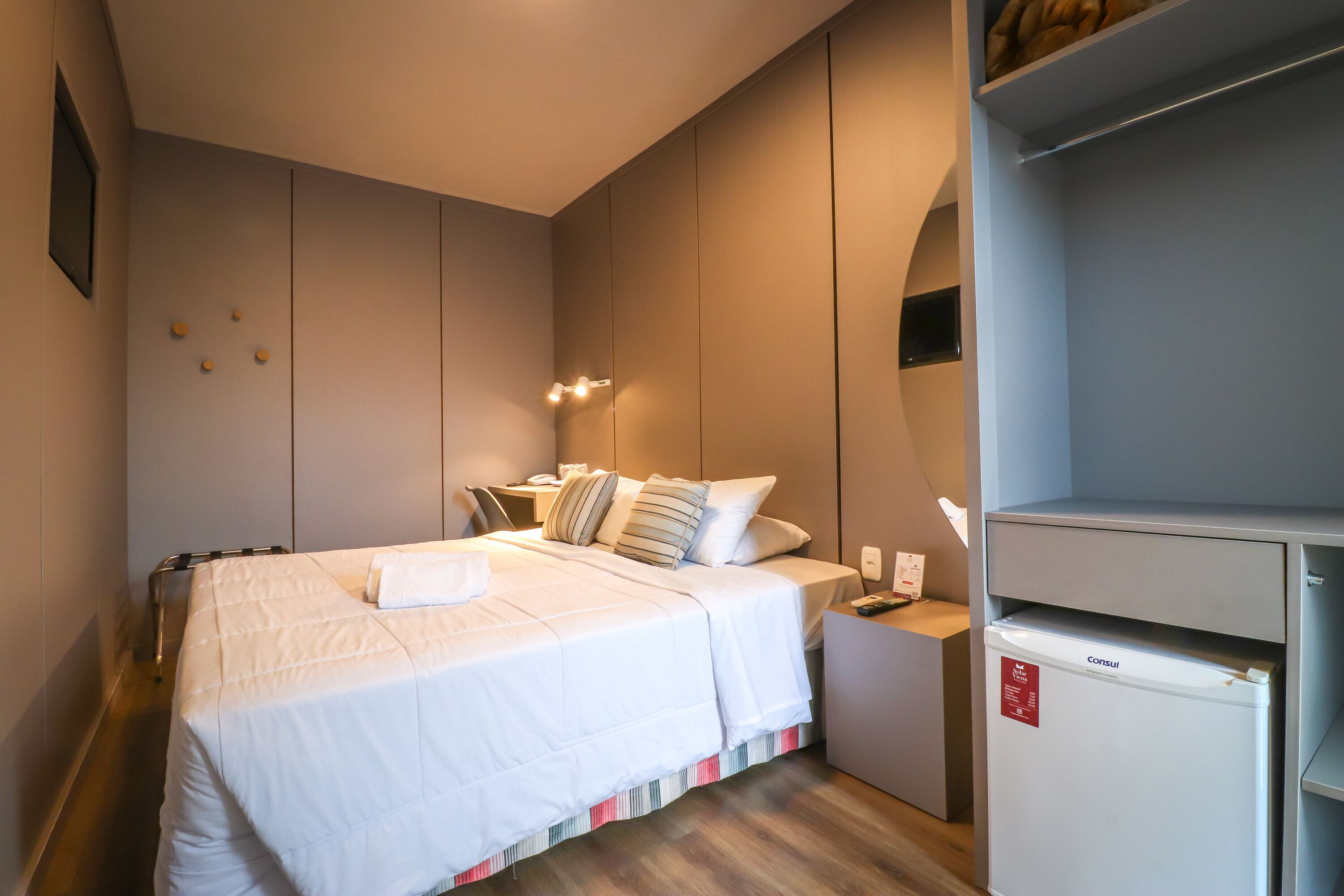 Quarto de hotel moderno com cama, mesa de cabeceira e minibar.