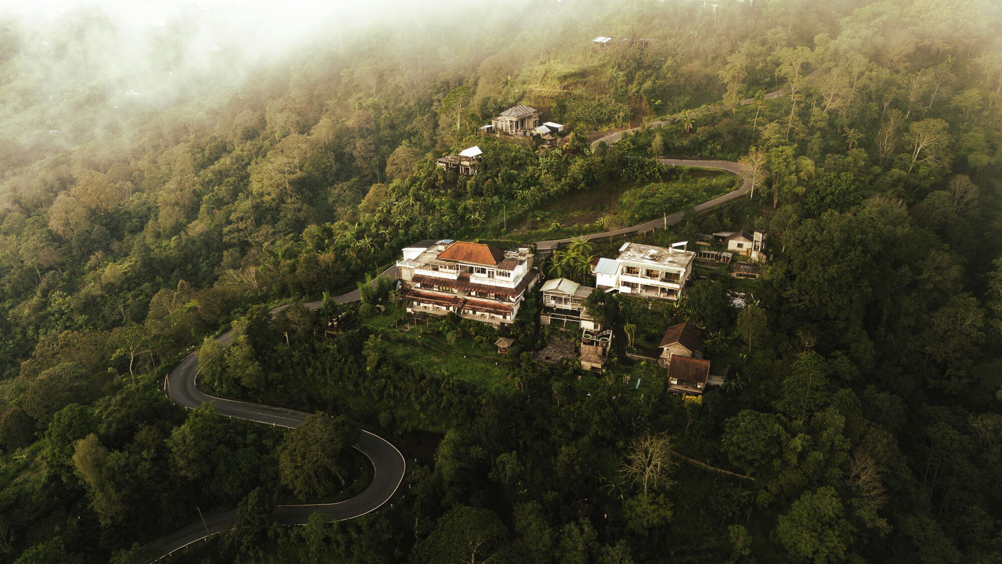 Vista aérea de uma propriedade em encosta com vários edifícios, uma estrada sinuosa e floresta densa.