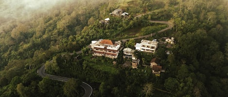 Vista aérea de uma propriedade em encosta com vários edifícios, uma estrada sinuosa e floresta densa.