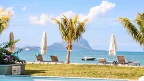Área da piscina com espreguiçadeiras e guarda-sóis, uma palmeira e vista para o mar com barcos e uma ilha ao longe.