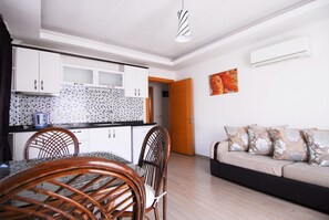 Uma cozinha moderna com armários brancos e painel de azulejos, uma sala de jantar com mesa de vidro e cadeiras de vime, e uma sala de estar com sofá e almofadas decorativas.