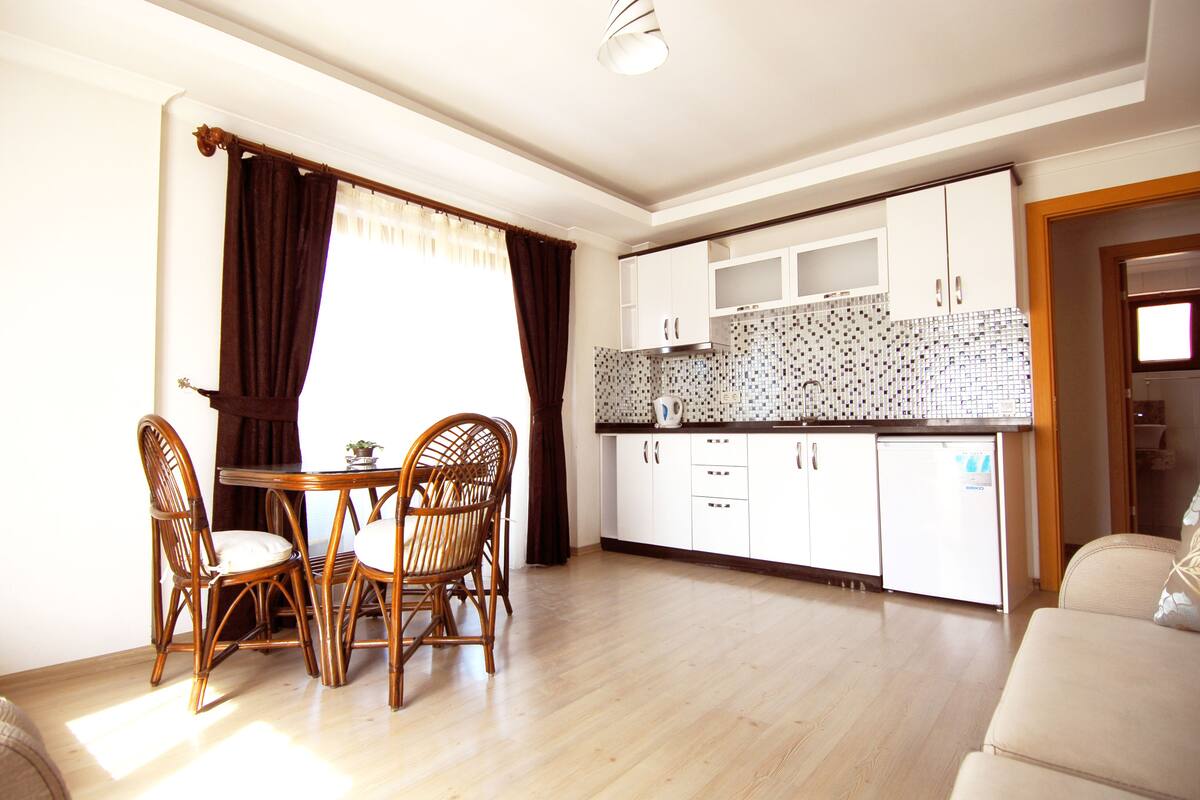 Uma sala de jantar com mesa e cadeiras, uma cozinha com armários brancos e painel de azulejos, e uma porta que dá para outro cômodo.