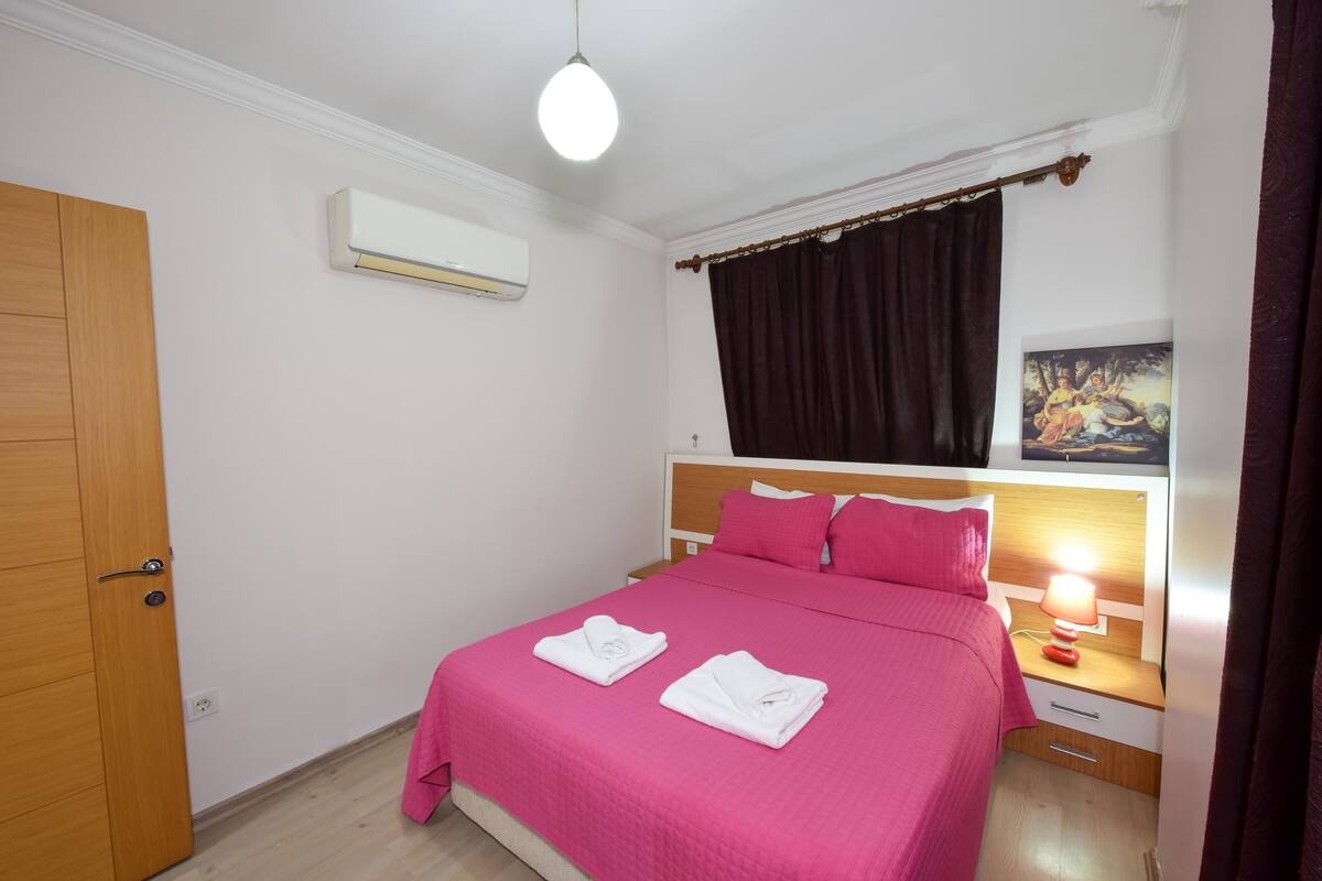 Quarto com cama, mesa de cabeceira, aparelho de ar condicionado, porta e um quadro na parede.