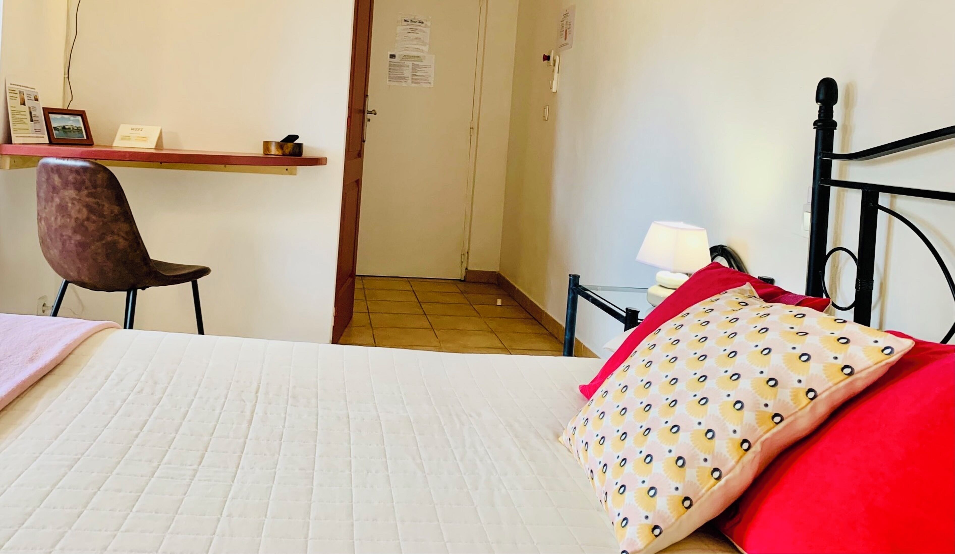 Quarto de hotel com cama, uma cadeira, escrivaninha e porta que dá para outro espaço.
