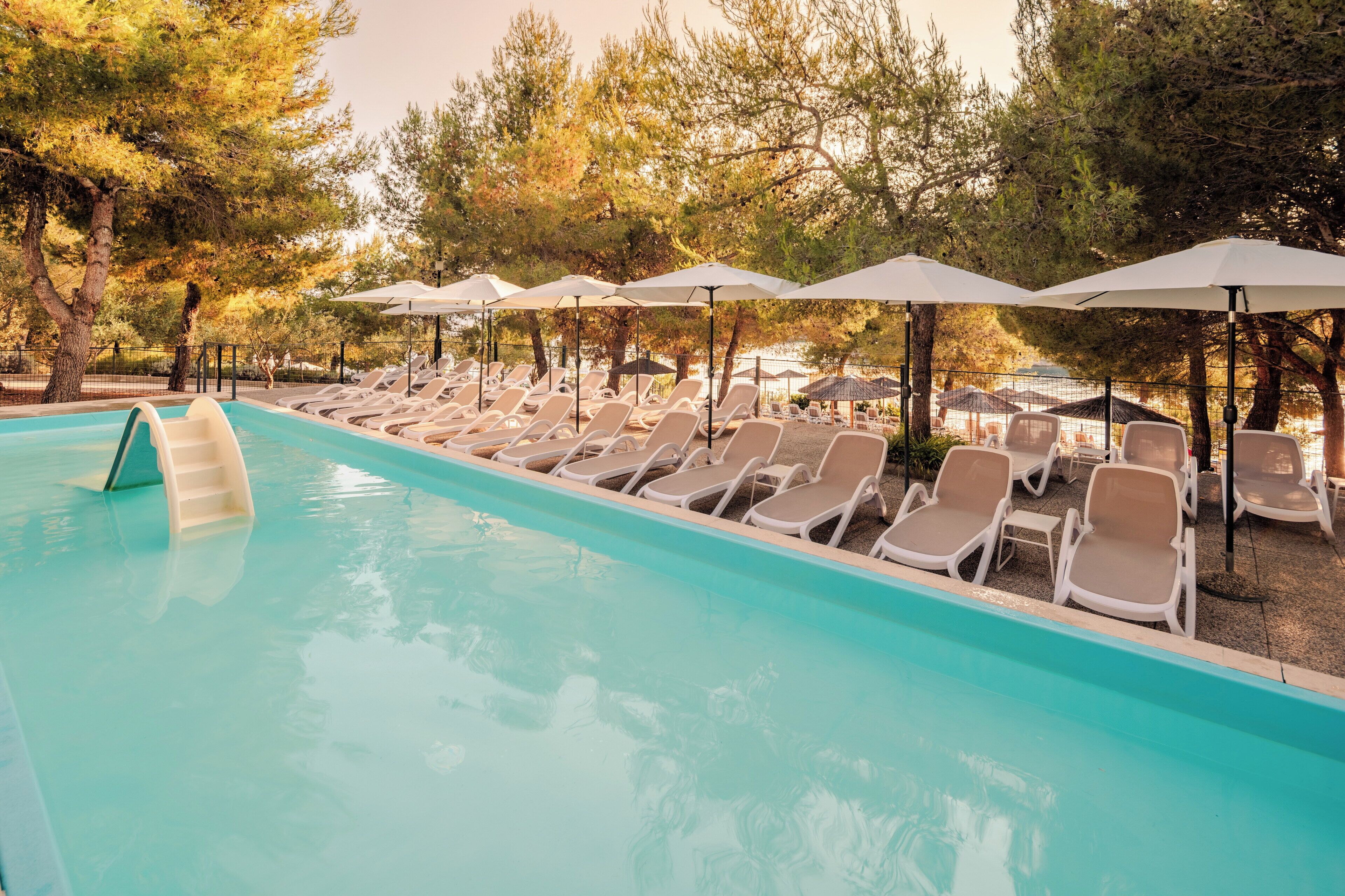 Uma área junto à piscina com espreguiçadeiras e guarda-sóis, rodeada por árvores.