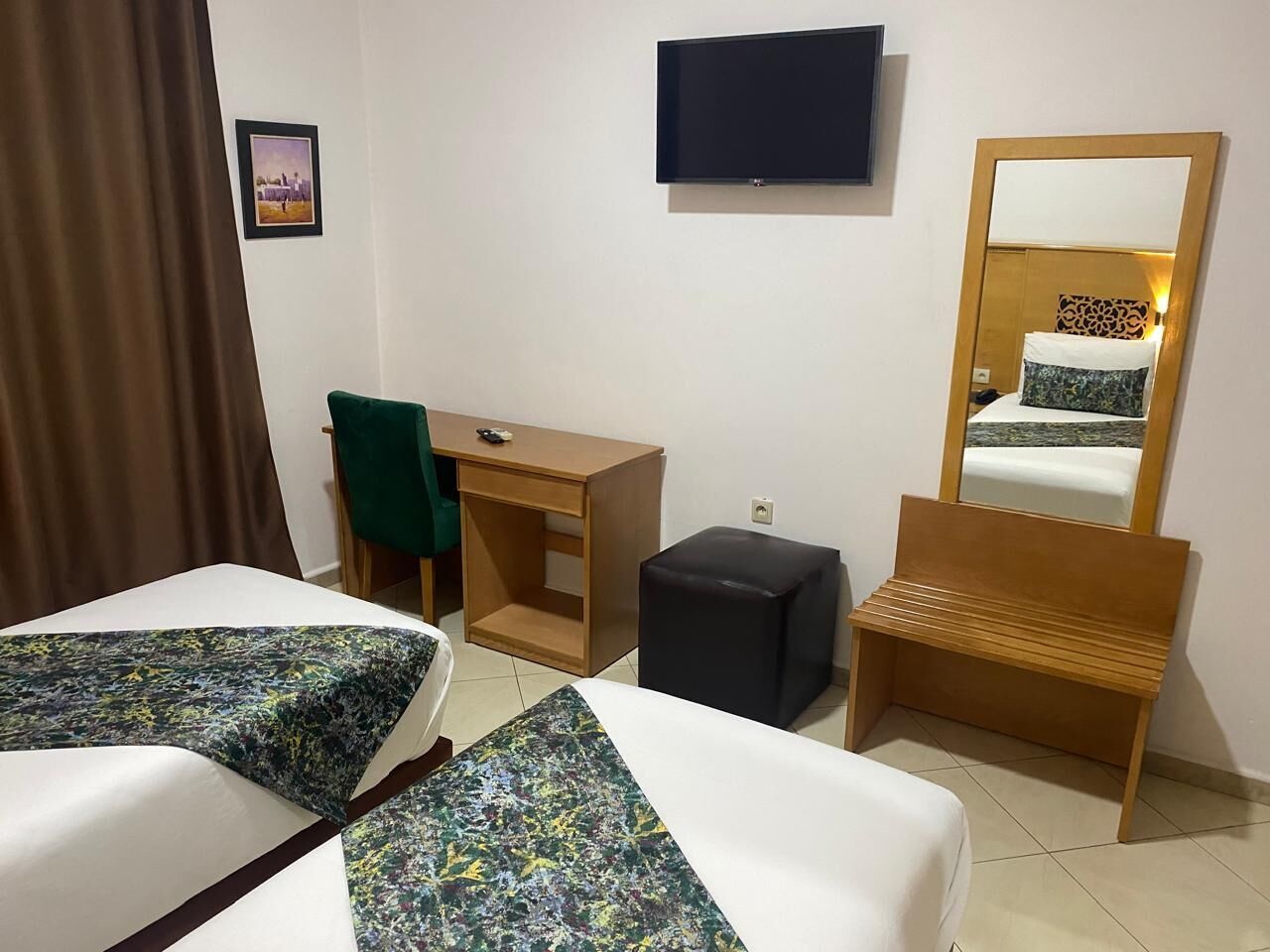 Quarto de hotel com duas camas, uma escrivaninha, uma cadeira, um pequeno banquinho, uma televisão e um espelho.
