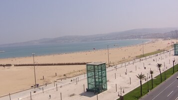 Uma praia com vista clara para o mar, um calçadão com estrutura de vidro e uma estrada ao lado.