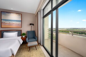 Quarto de hotel com cama, uma cadeira, mesa de cabeceira com abajur, pintura de uma praia e varanda com vista para a cidade.