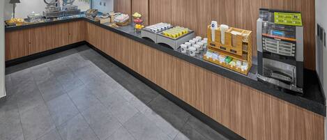 Uma zona de pequeno-almoço moderna com uma máquina de café, cestos de fruta e diversas opções de pequeno-almoço numa bancada de madeira.